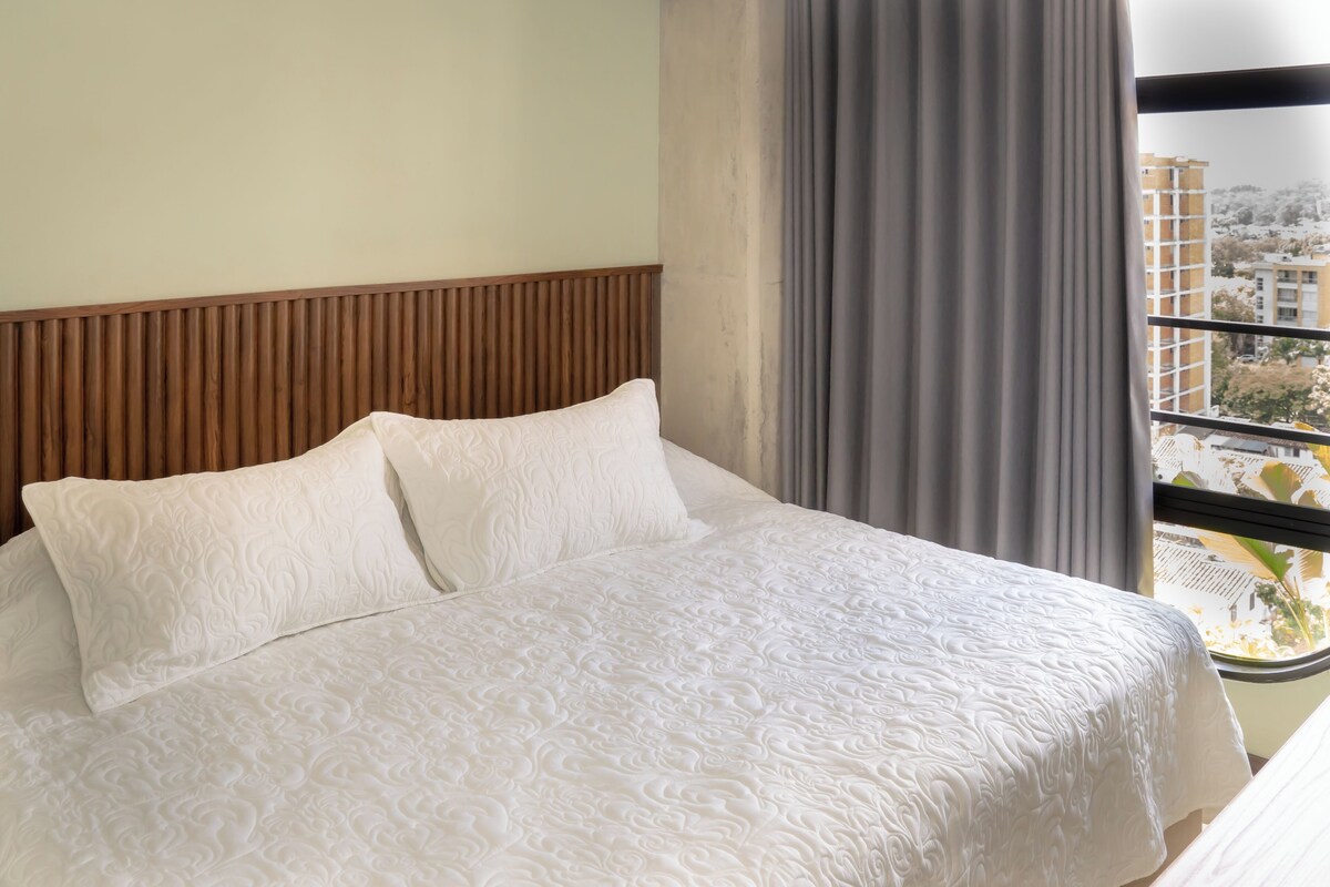 Quarto de hotel com cama, cabeceira de madeira e janela com vista para edifícios.