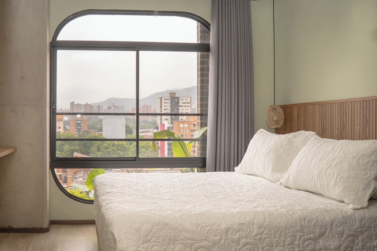 Quarto com uma grande janela, cama com roupa de cama branca e vista para edifícios e vegetação.