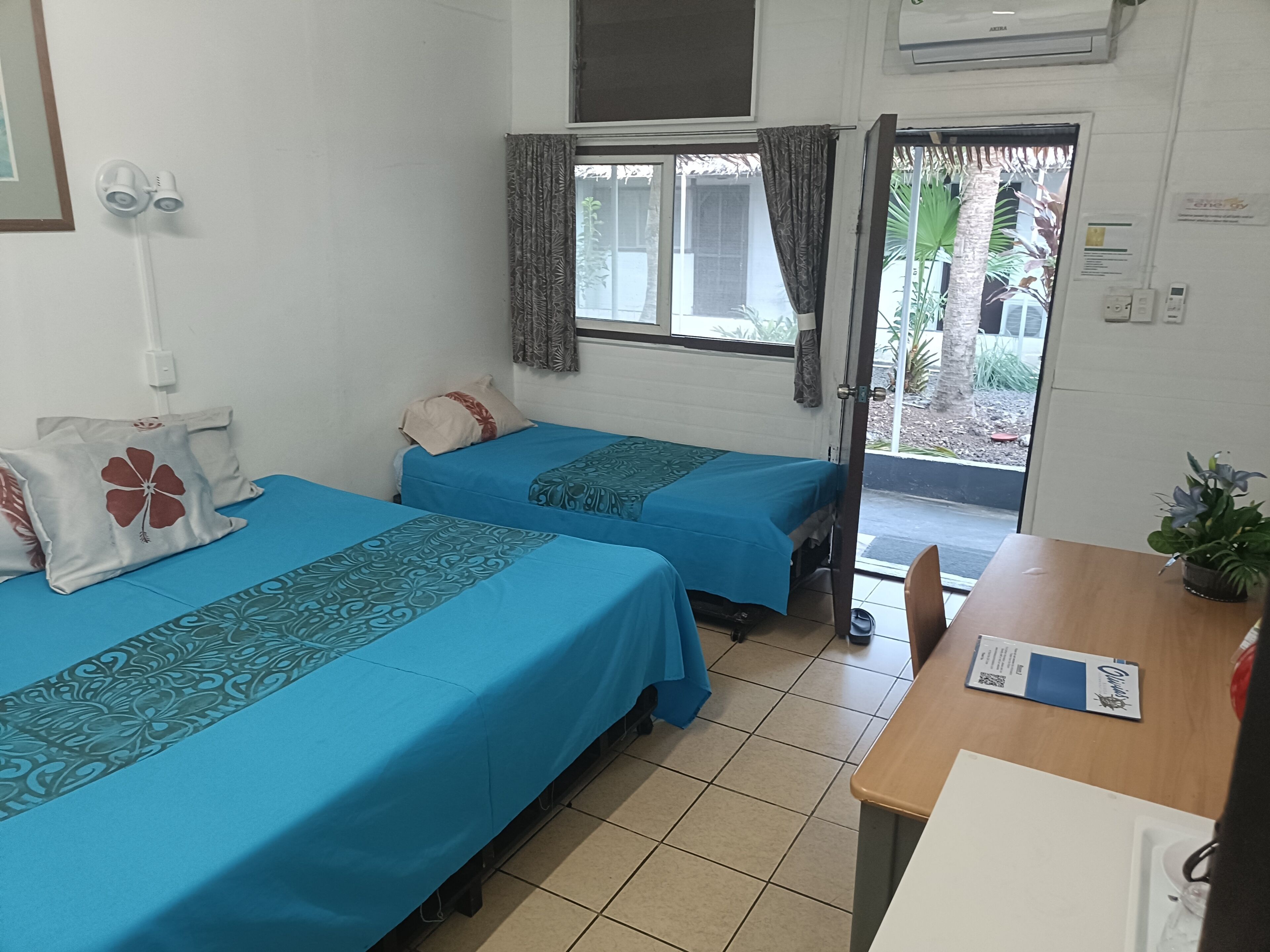 Quarto com duas camas, uma mesa pequena, uma janela com cortinas e um aparelho de ar condicionado.