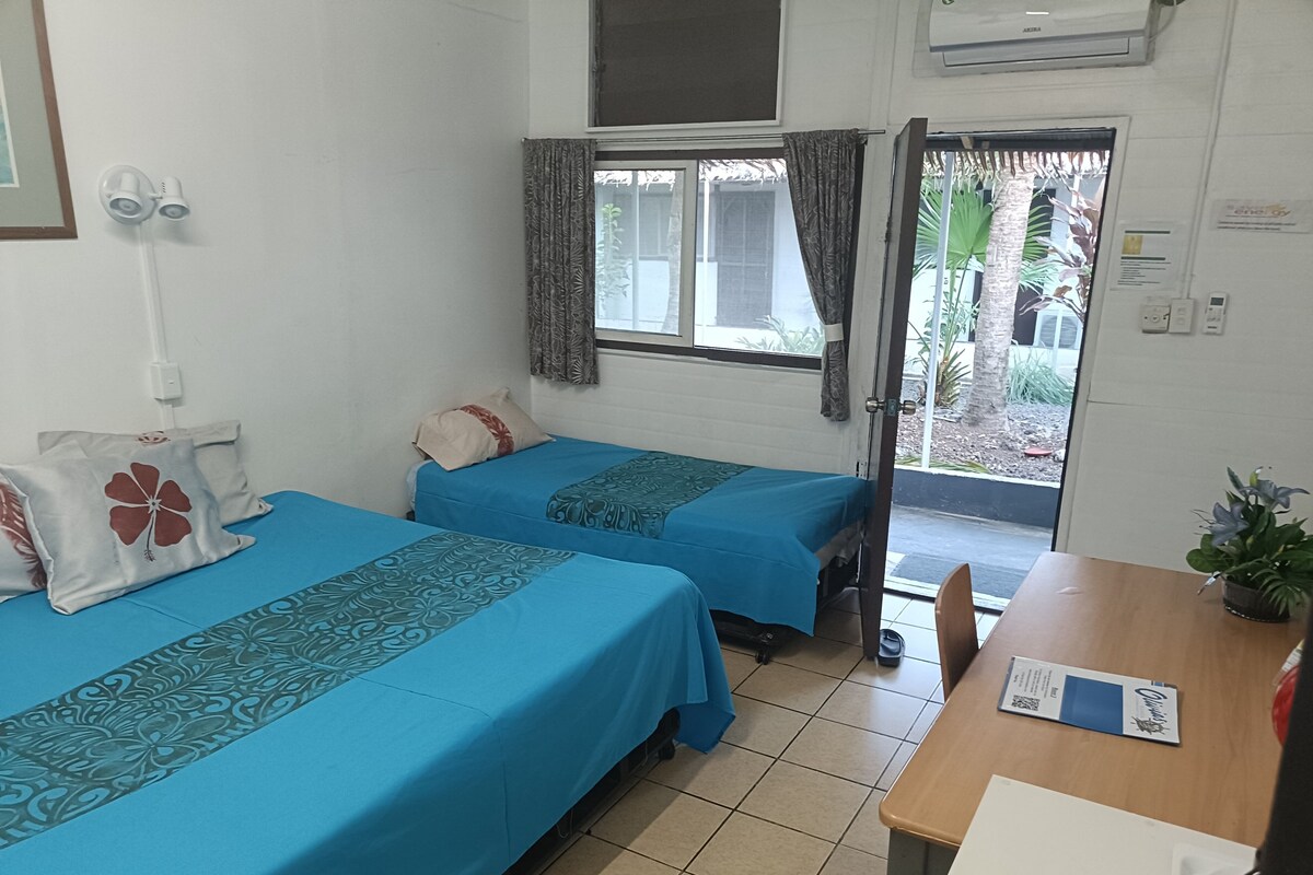 Quarto com duas camas, uma mesa pequena, uma janela com cortinas e um aparelho de ar condicionado.