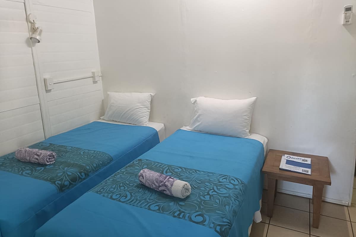 Quarto pequeno e limpo com duas camas, uma mesa de cabeceira de madeira e uma porta branca.