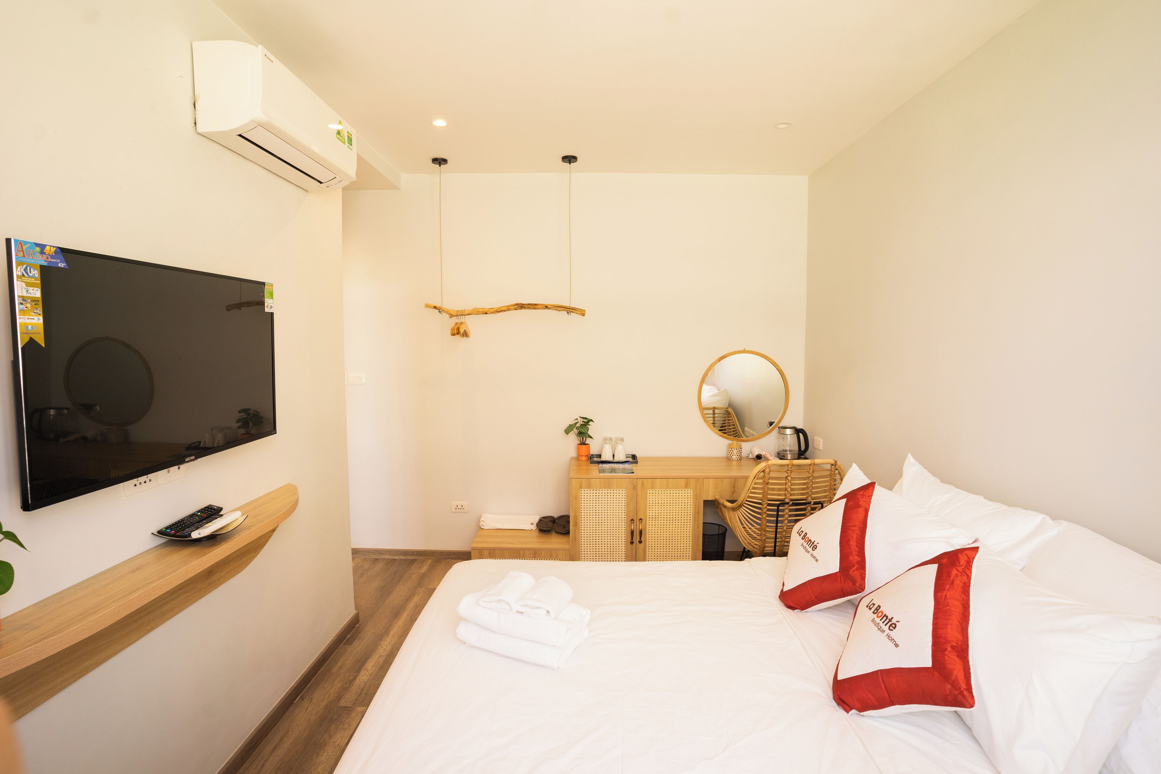 Quarto de hotel com televisão de tela plana, mesa de cabeceira de madeira, cama com lençóis brancos e almofadas vermelhas, cômoda de madeira, cadeira de vime e um espelho.