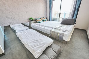 Quarto moderno com uma cama grande, uma cama menor e um sofá. Há uma planta em uma mesinha de cabeceira e uma janela com cortinas.