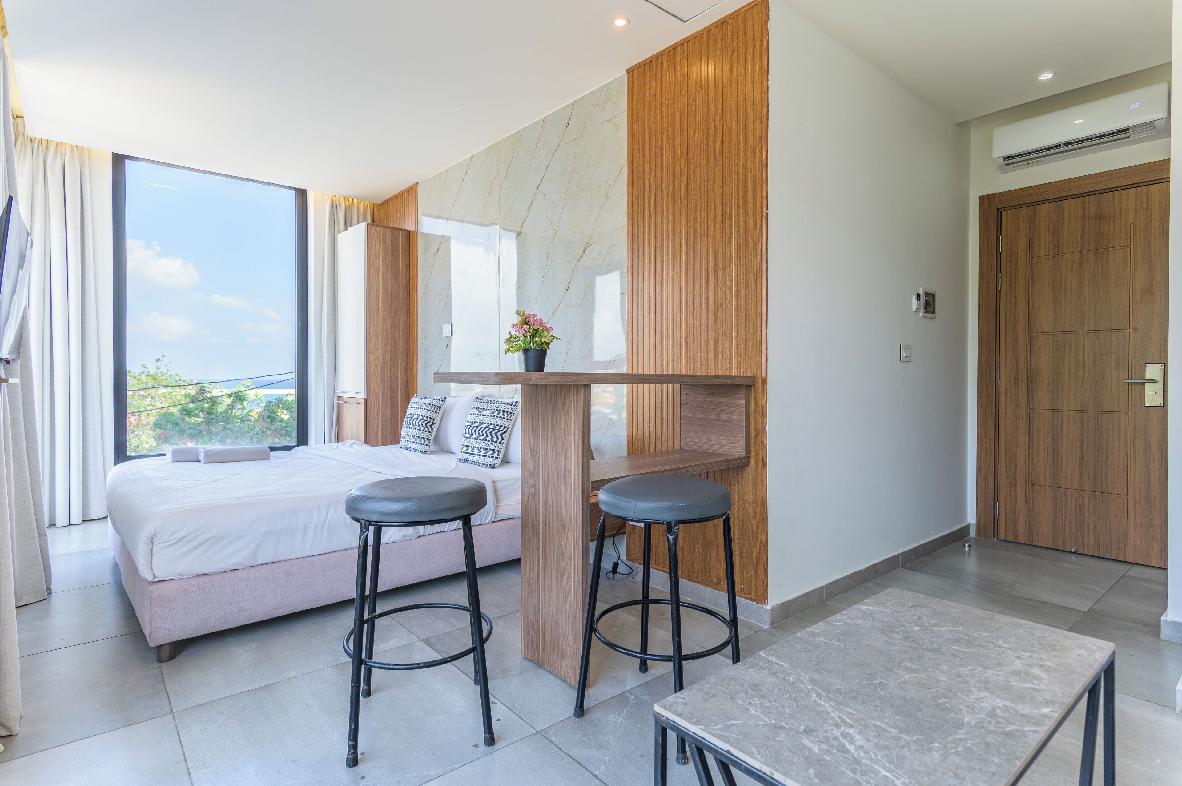 Quarto moderno com uma cama grande, dois banquinhos, uma mesinha e vista para o exterior.