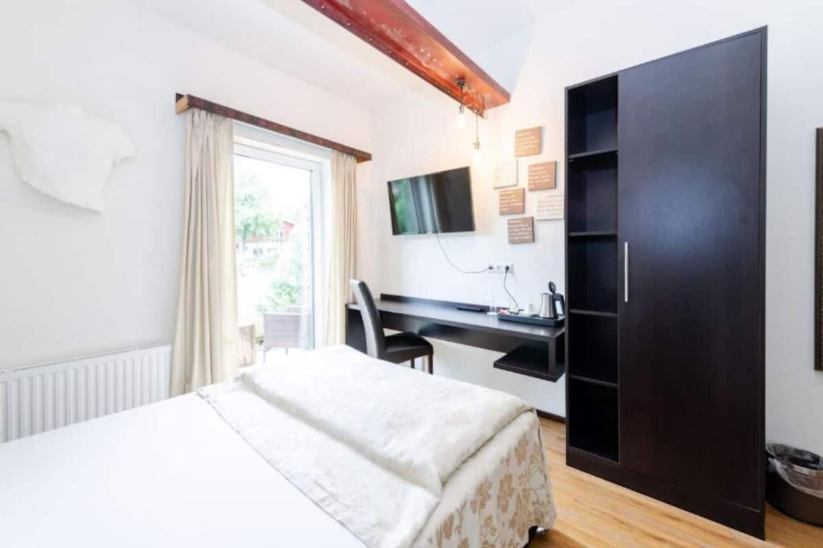 Quarto de hotel com cama, escrivaninha, cadeira, guarda-roupa, televisão e janela com cortinas.