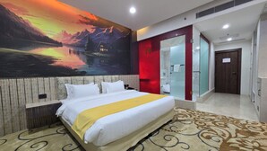 Quarto de hotel com cama grande, parede de destaque vermelha, box de chuveiro em vidro e um mural com pôr do sol sobre montanhas.