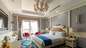 Quarto de hotel com uma cama grande, uma cadeira vermelha, uma mesa com abajur e um candelabro.