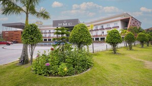 Um hotel com design moderno, exibindo uma placa HMC em destaque, jardins ajardinados e estacionamento.