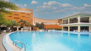 Uma piscina grande e límpida com borda curva, rodeada por palmeiras e edifícios modernos.