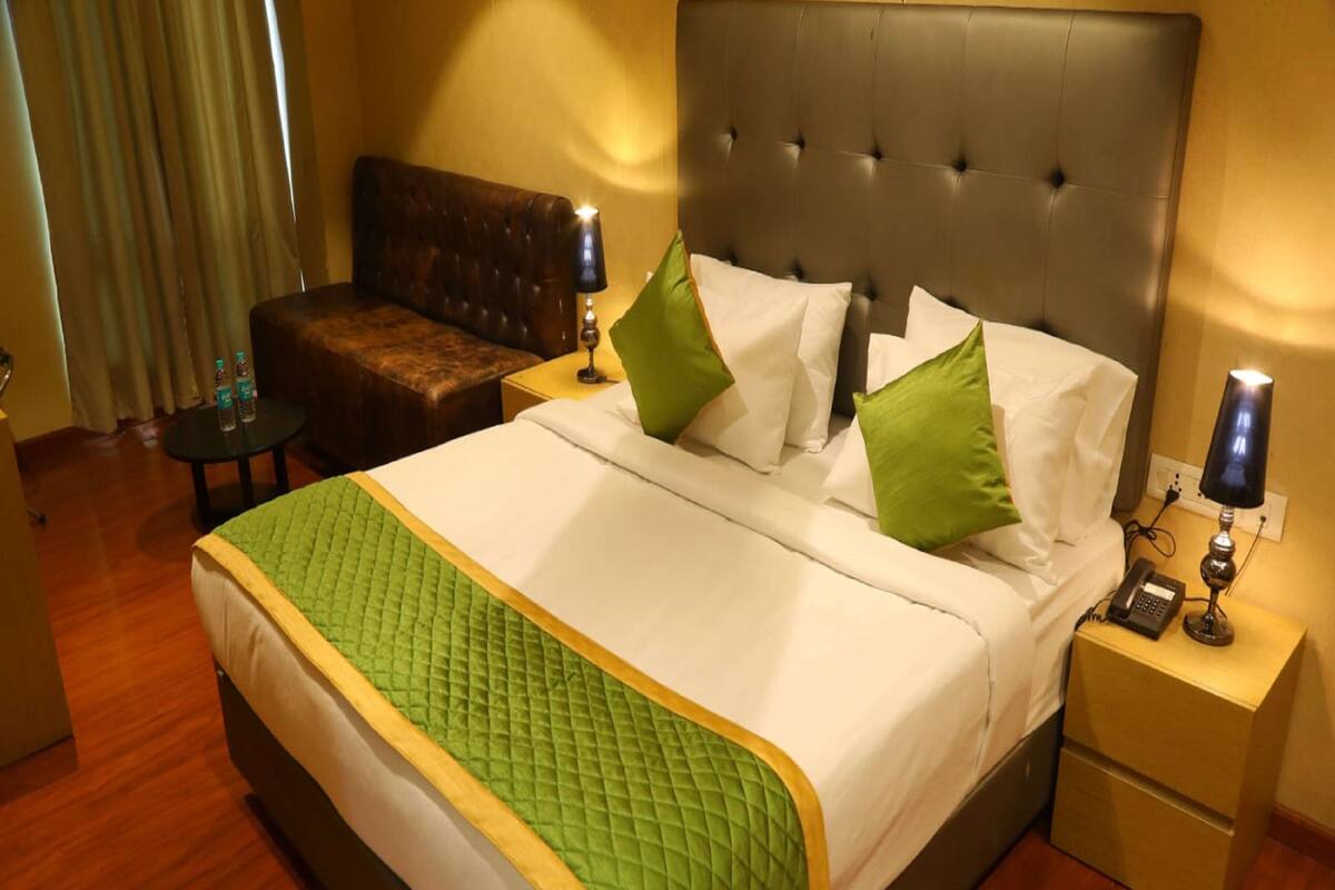 Quarto de hotel com uma cama grande, cabeceira, duas mesas de cabeceira, um banquinho e um edredom acolchoado verde, além de um otomano de couro marrom.
