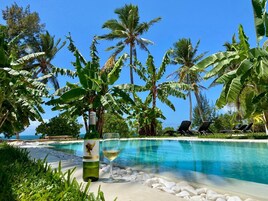 Uma área à beira da piscina com uma garrafa de vinho e um copo, cercada por vegetação tropical exuberante e palmeiras.