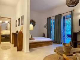 Quarto com cama, mesa de cabeceira de madeira, espelho e vista para o exterior através de uma porta de correr.