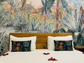 Cama com roupa de cama branca, duas almofadas com motivos tropicais e um mural de uma cena de selva na parede.