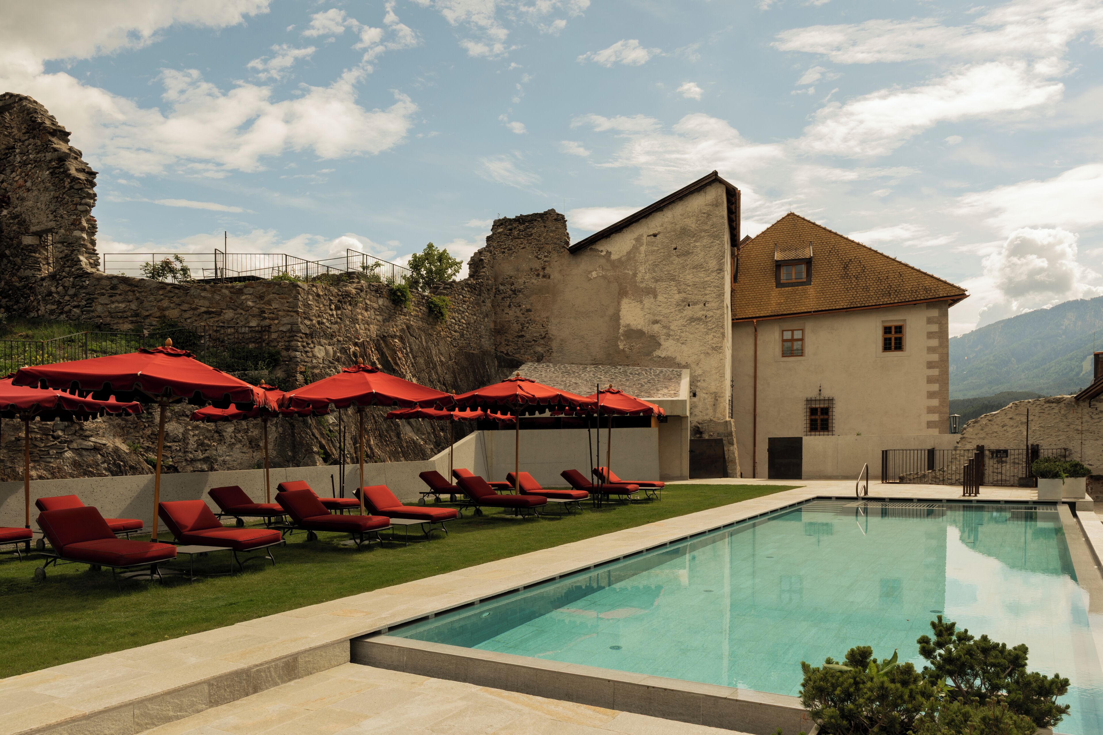 Piscina com guarda-sóis vermelhos, espreguiçadeiras e um edifício com parede de pedra e vista para a montanha.