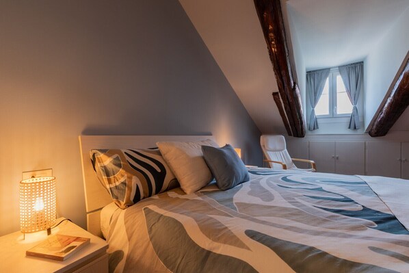 Quarto com cama, uma cadeira, mesa de cabeceira e janela com cortinas.