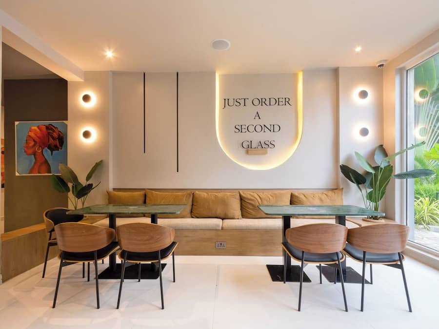 Uma zona de refeições moderna com mesa de vidro, cadeiras de madeira, um sofá e uma parede com luminária circular exibindo o texto "JUST ORDER A SECOND GLASS".