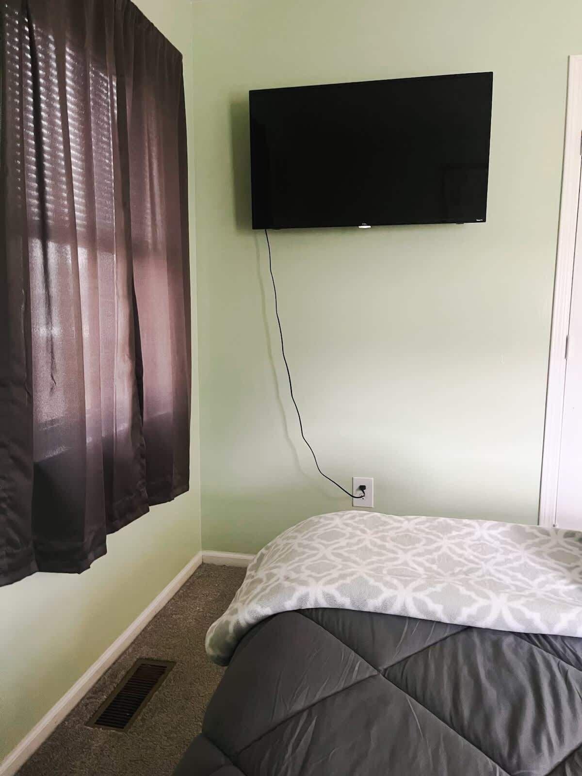 Quarto com cama, televisão na parede, tomada e janela com cortinas.