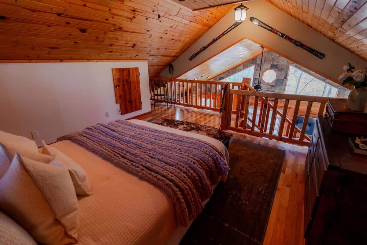 Um quarto de sótão acolhedor com teto de madeira, uma cama com manta roxa e guarda-corrimão de madeira, com vista para o exterior.