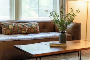 Sala de estar com um sofá de couro marrom, almofada floral, mesa de centro de madeira e um vaso com plantas verdes.