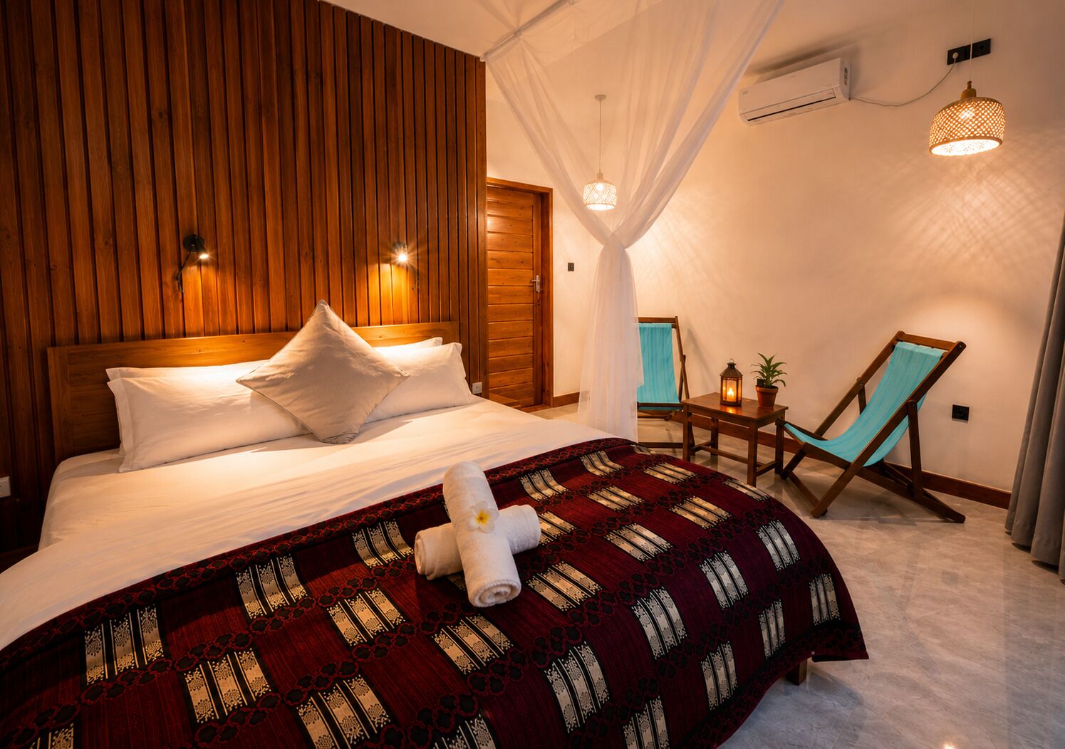 Quarto de hotel com cabeceira de madeira, cama com roupa de cama branca, colcha de cama vermelha estampada, uma cadeira azul e uma pequena mesa com abajur.