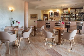 Área de bar de hotel com mesas e cadeiras redondas, balcão de bar com espelho e uma parede de garrafas de bebidas.