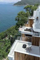 Um edifício moderno com varanda de vidro com vista para um corpo de água e uma encosta arborizada.