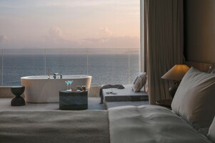 Quarto de hotel moderno com uma banheira grande, um sofá, uma mesa pequena e vista para o mar.