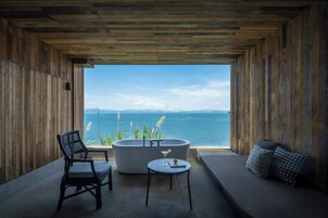 Quarto com piso de madeira, banheira grande, sofá e poltrona, com vista para um corpo d'água e montanhas ao longe.