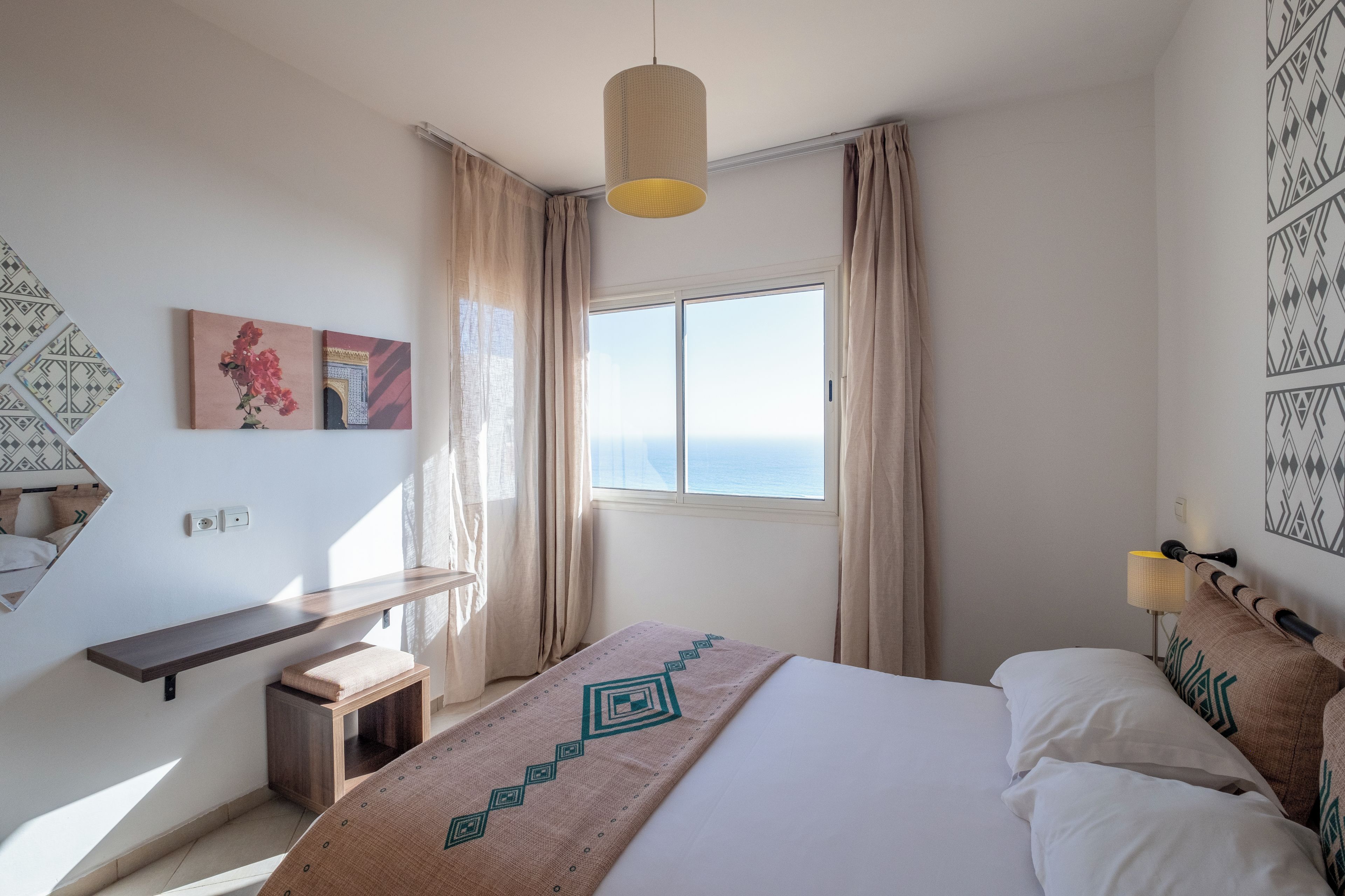 Quarto de hotel com cama, mesa de cabeceira, janela com cortinas, luminária na parede e um quadro.