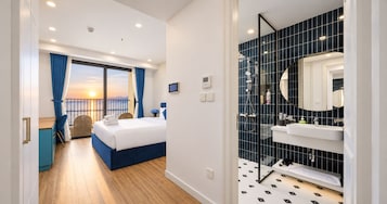 Quarto de hotel com cama, escrivaninha, cadeira, varanda com cortinas e banheiro com espelho e pia.