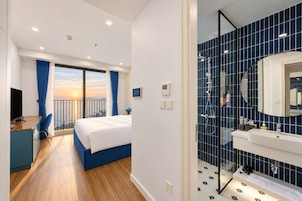Quarto de hotel com varanda, cama, escrivaninha e banheiro com espelho grande e azulejos azuis.