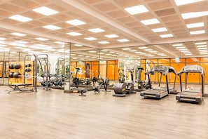 Academia com diversos equipamentos de ginástica, paredes com espelhos e piso de madeira.
