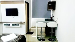 Quarto de hotel compacto com cama, televisão presa à parede, escrivaninha com computador e dois bancos altos brancos.