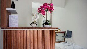 Uma receção com bancada de madeira, duas orquídeas em vasos, uma placa emoldurada e uma cadeira moderna.