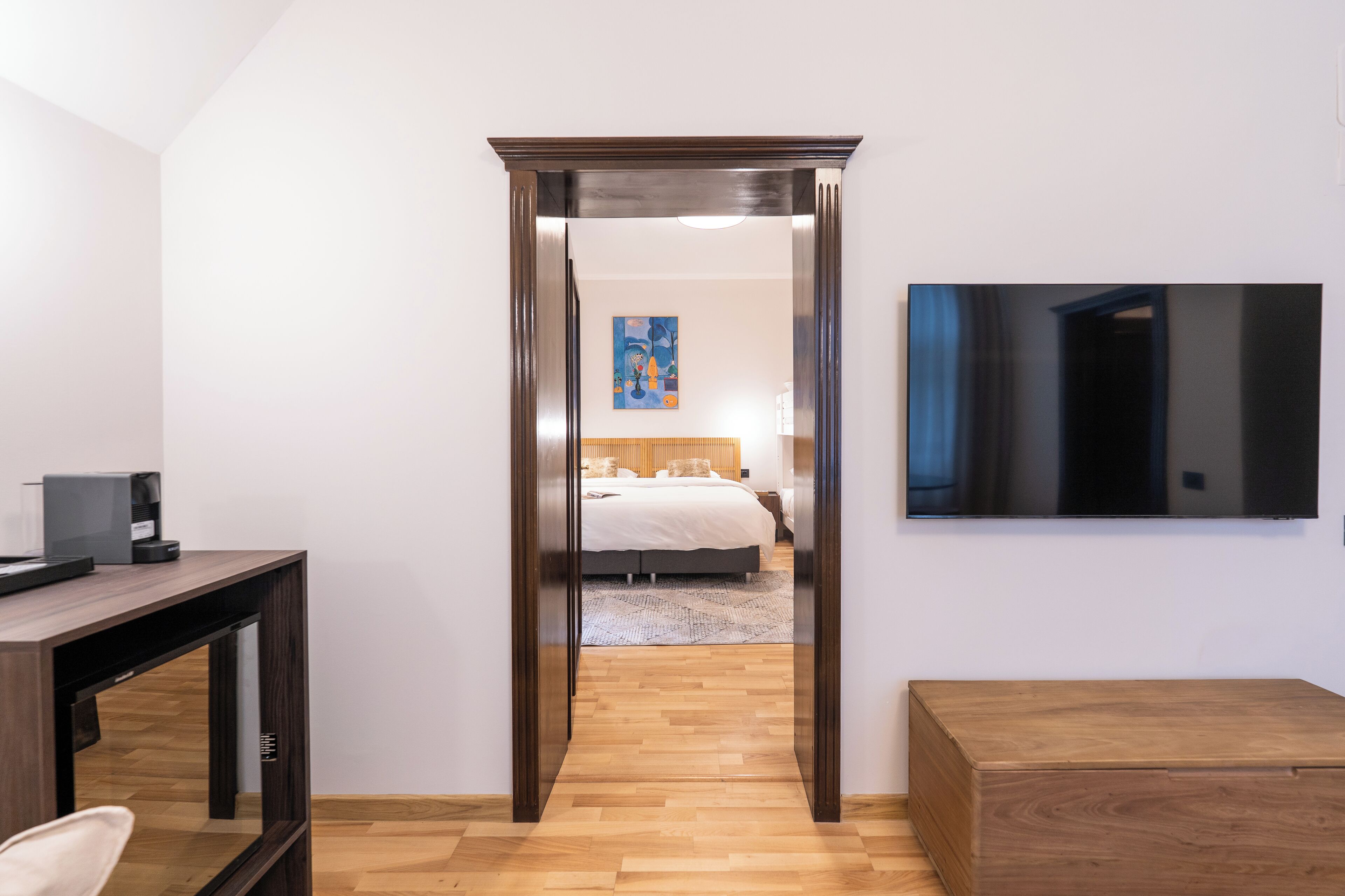 Quarto de hotel com piso de madeira, cama com roupa de cama branca, TV de tela plana na parede e máquina de café sobre uma mesa de madeira.