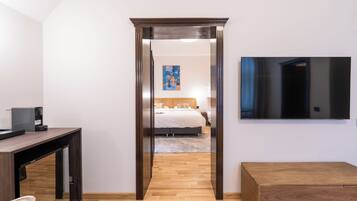 Quarto de hotel com piso de madeira, cama com roupa de cama branca, TV de tela plana na parede e máquina de café sobre uma mesa de madeira.