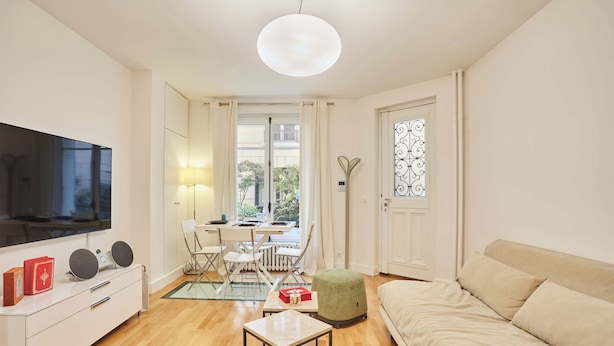 Uma sala de estar moderna com televisão de tela plana, uma área de jantar com mesa e cadeiras, um sofá e um pequeno banquinho.