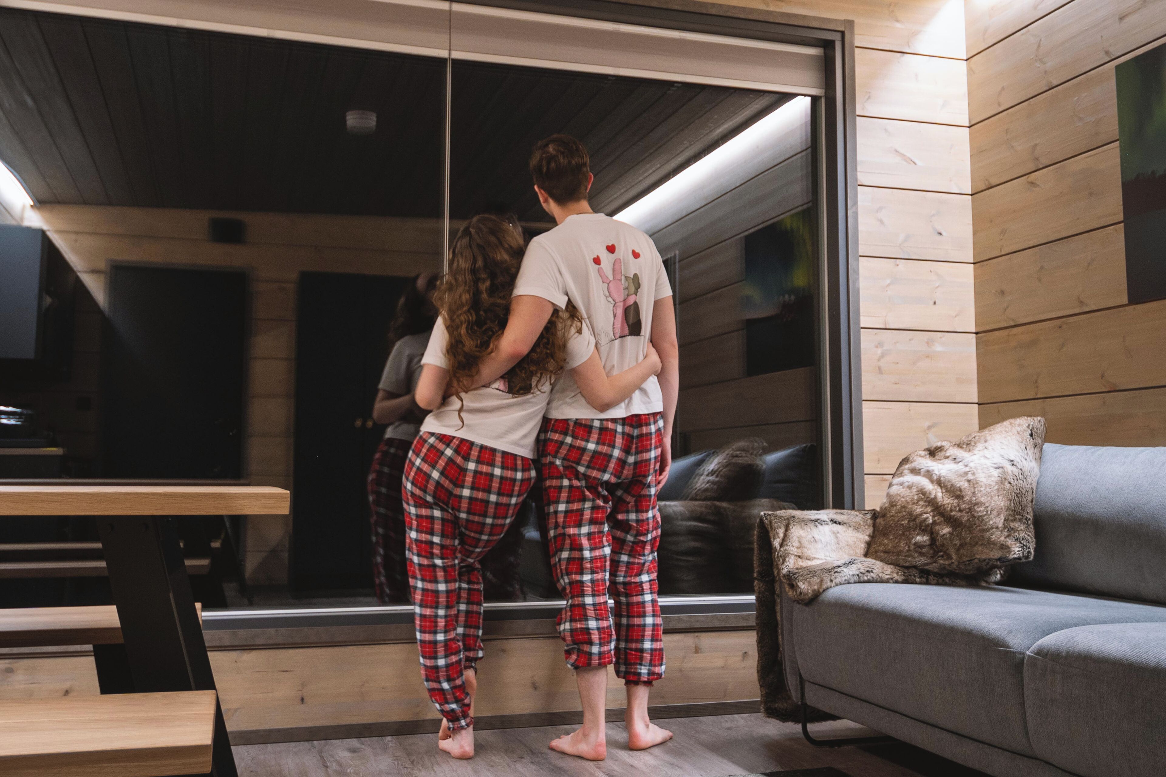 Um casal em um quarto moderno com um espelho grande, paredes de madeira e um sofá cinza.