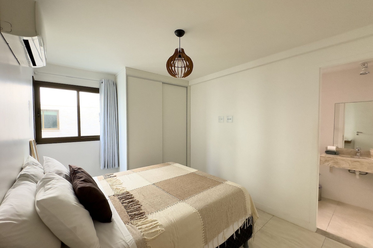 Quarto com cama, janela, guarda-roupa, luminária pendente e banheiro com espelho.
