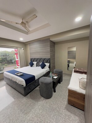 Quarto de hotel moderno com uma cama grande, uma escrivaninha, uma cadeira, um pequeno banquinho, um ventilador de teto e uma janela ampla com vista.