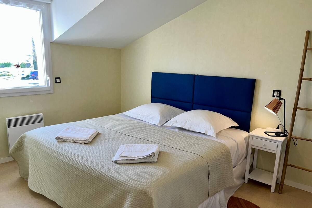Uma cama bem arrumada com cabeceira azul, abajur e janela com vista para o exterior.