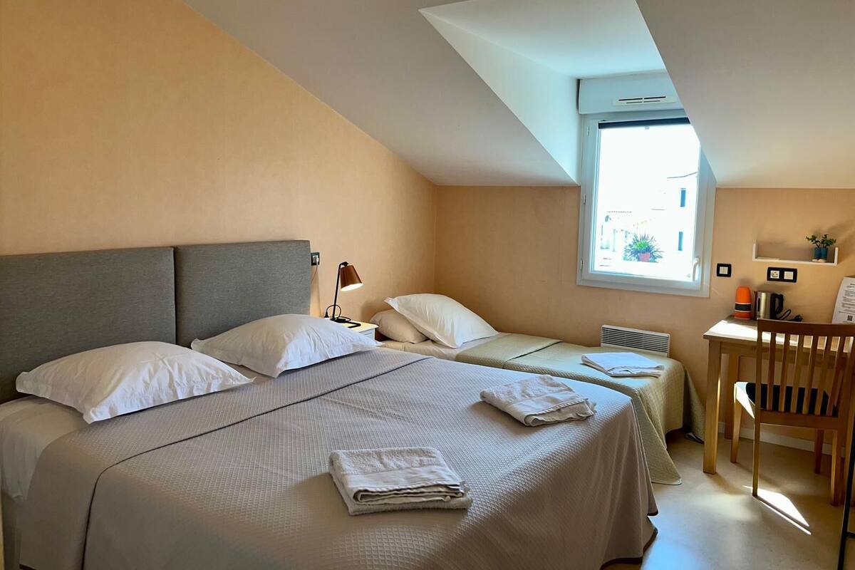 Quarto de hotel com duas camas, uma escrivaninha, uma cadeira, uma janela e um claraboia.