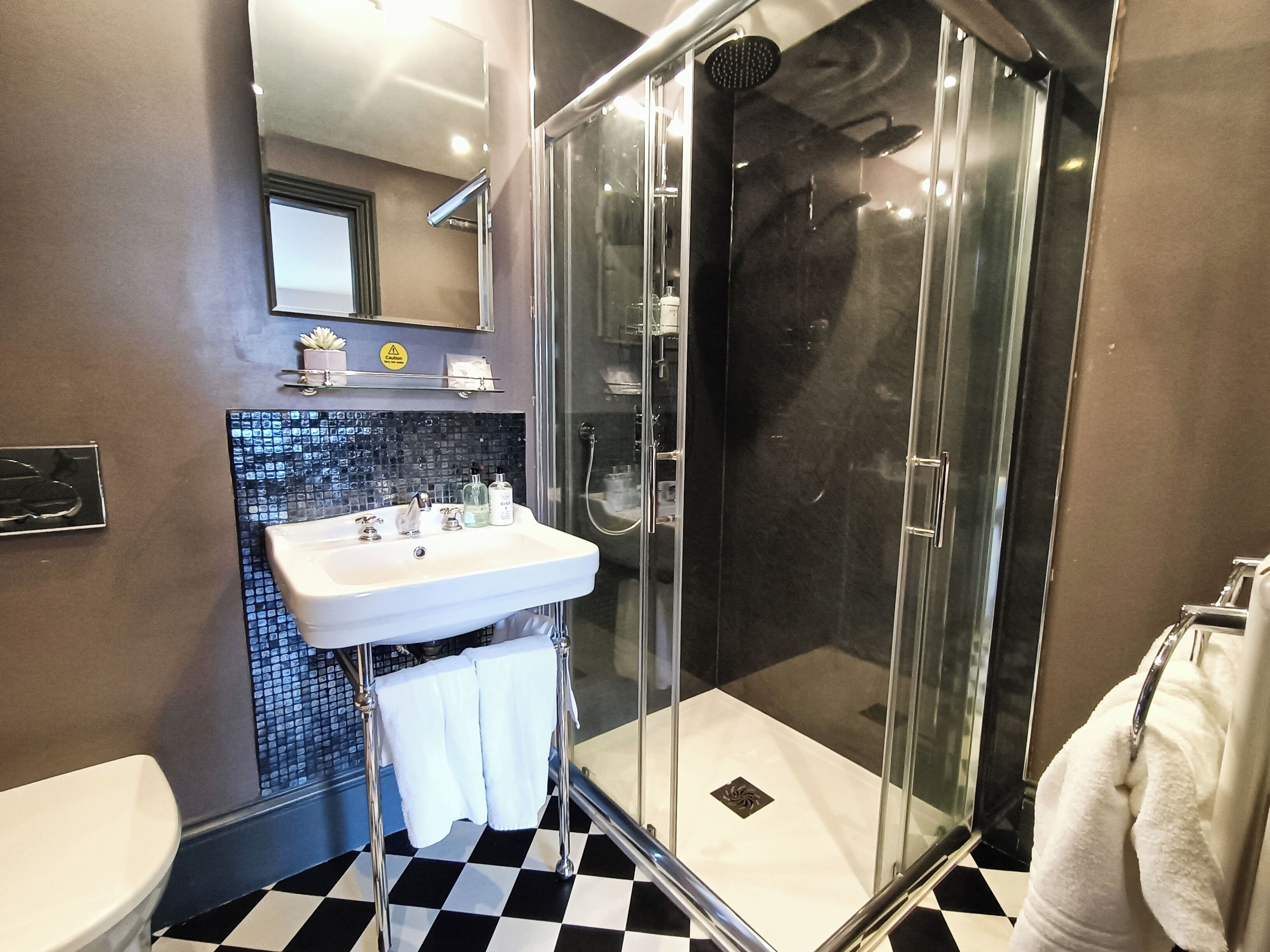 Casa de banho com chuveiro em cabine de vidro, lavatório branco com artigos de higiene e chão xadrez.