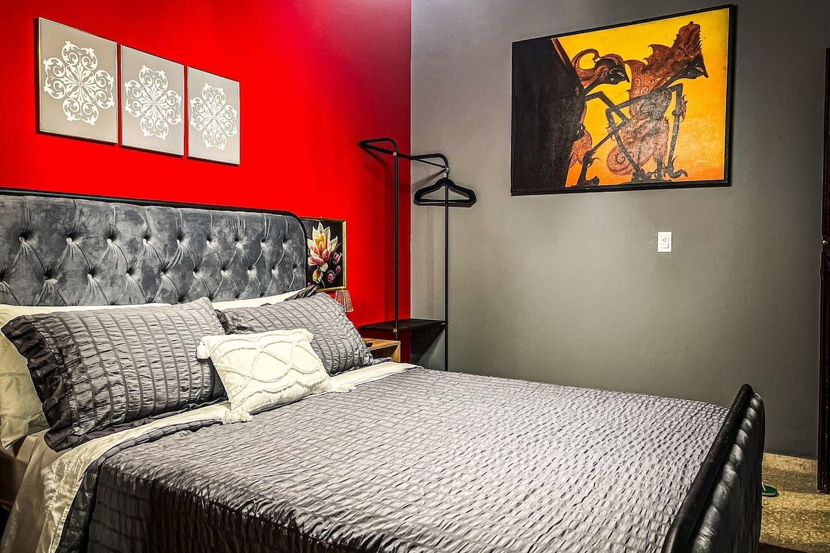Quarto com cabeceira estofada, cama com roupa de cama cinza, parede em destaque vermelha e três painéis decorativos brancos.