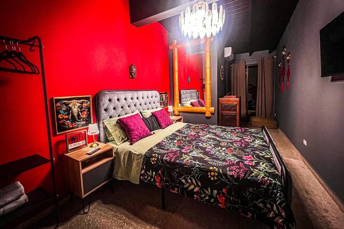 Quarto com cabeceira estofada, colcha floral, luminária de cabeceira e espelho fixado na parede.