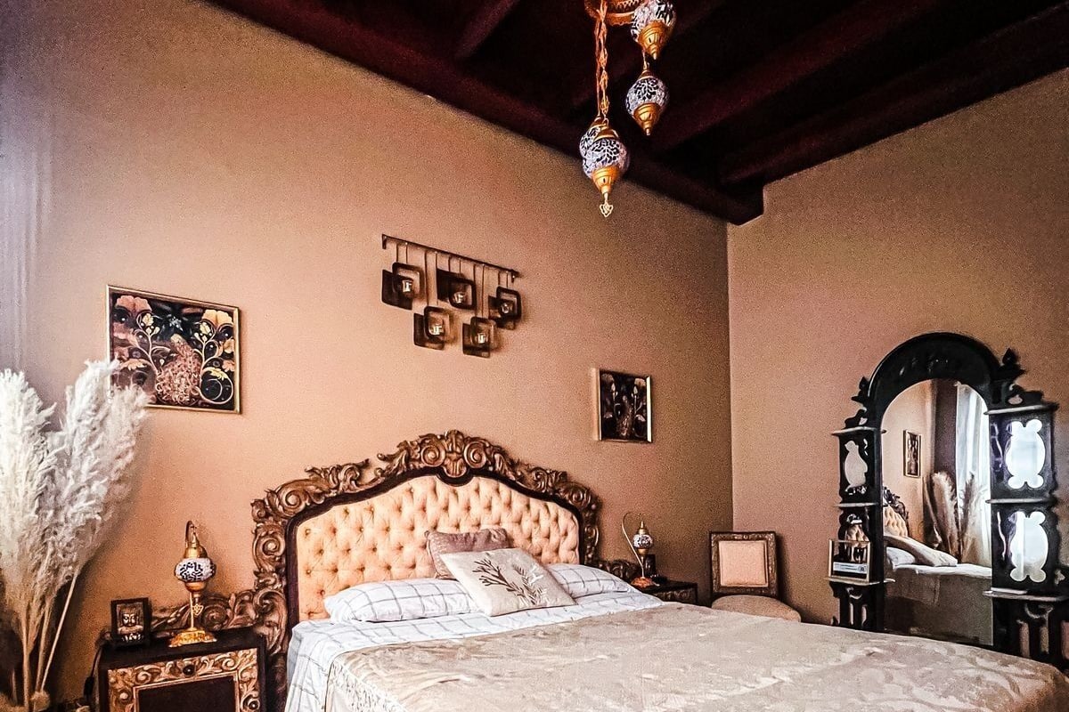 Quarto com uma cama grande, cabeceira ornamentada, arandelas decorativas e um lustre clássico.