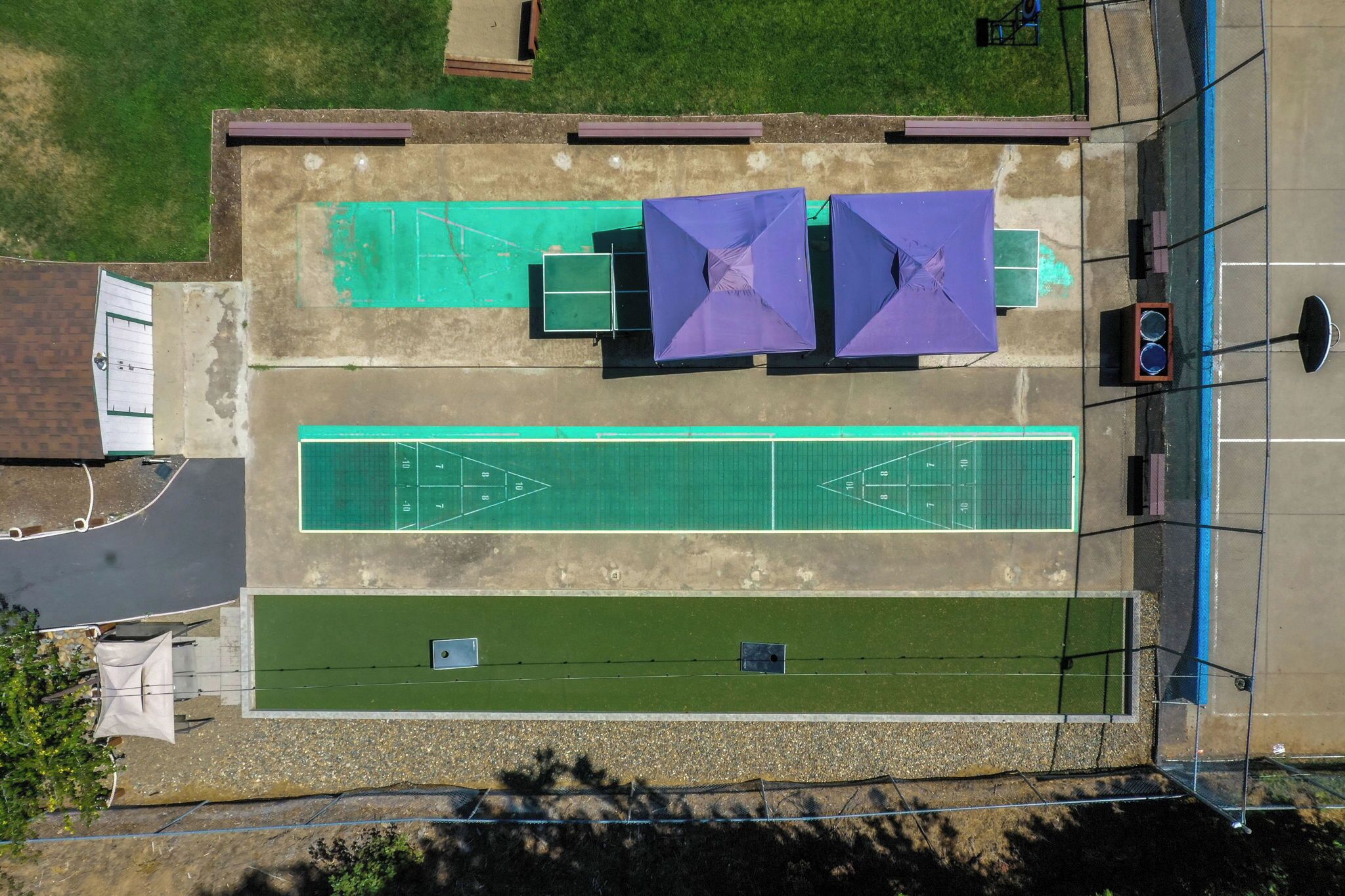Vista aérea de uma instalação esportiva com vários campos, incluindo uma quadra de basquete e uma quadra de tênis, cercada por uma grade e áreas sombreadas.