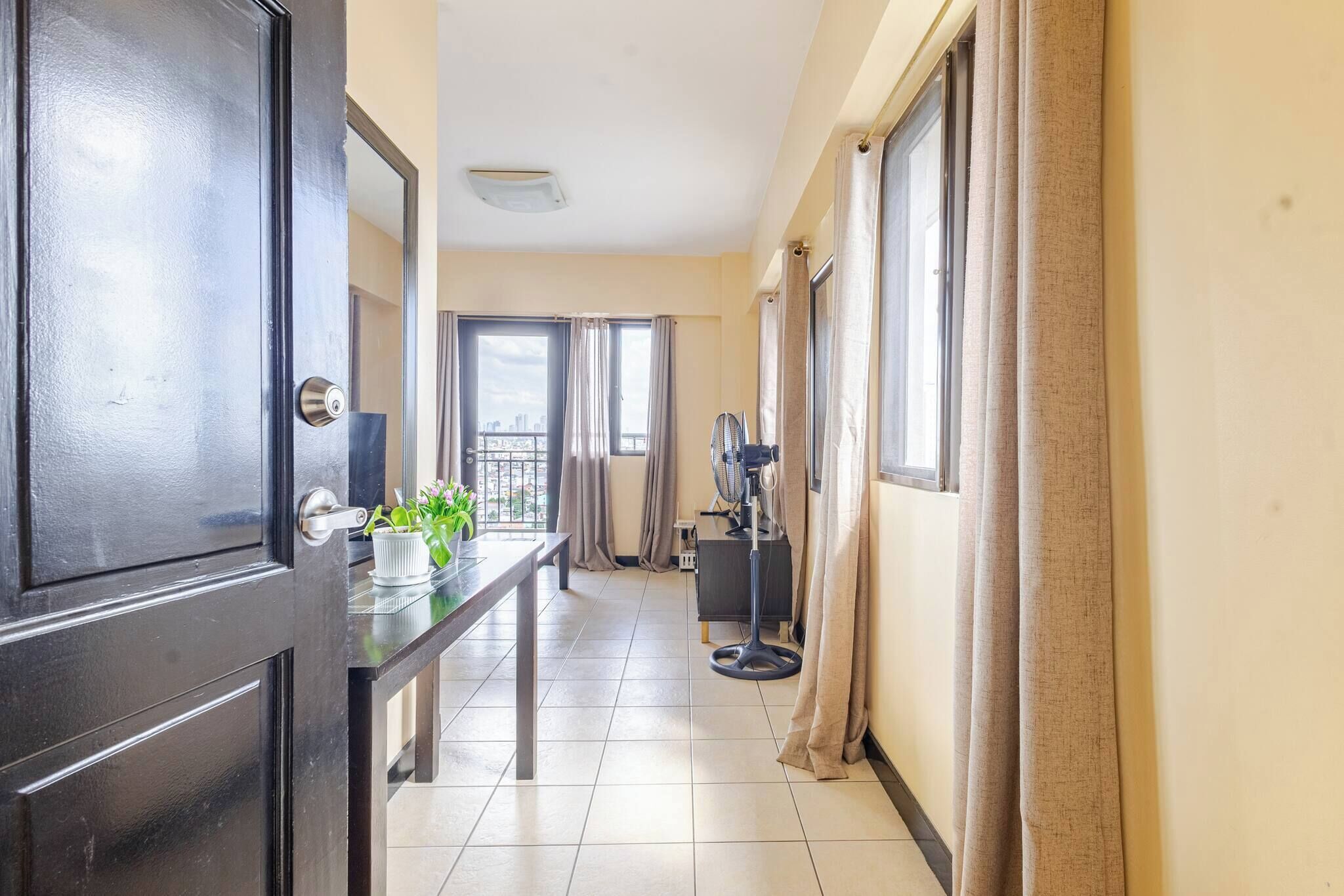 Um corredor com uma porta, uma mesa com uma planta, um ventilador de chão e uma janela com cortinas.