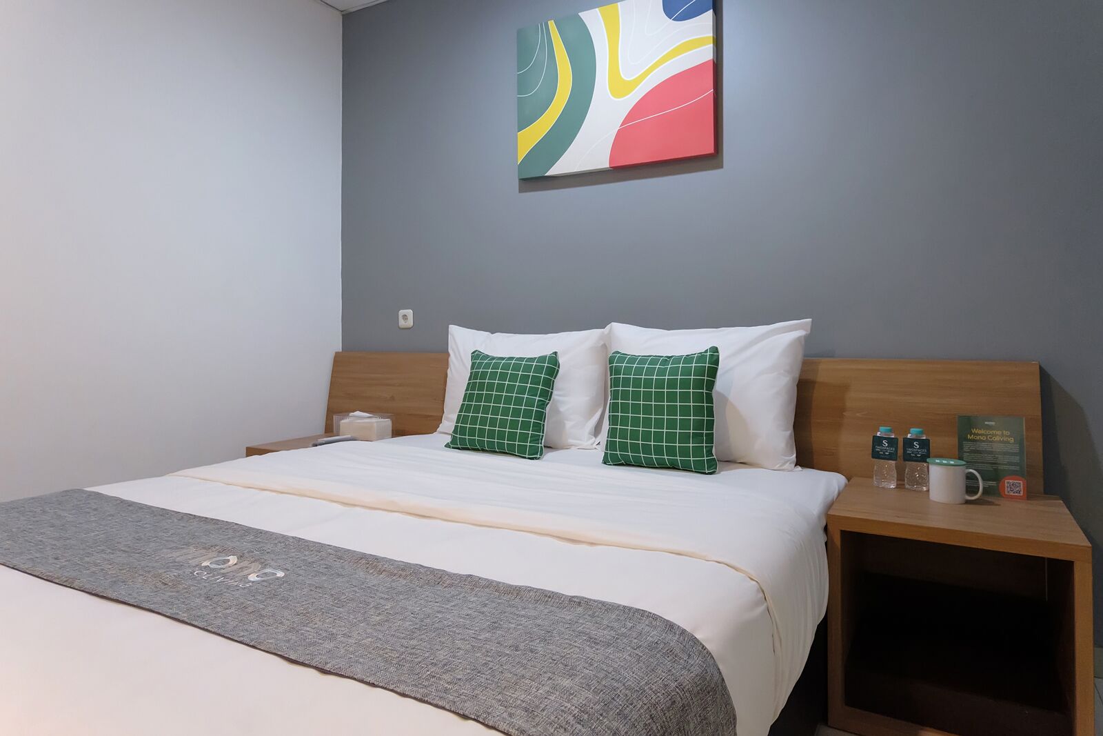Quarto de hotel com cama, duas almofadas, mesa de cabeceira, caneca de café e placa de boas-vindas.