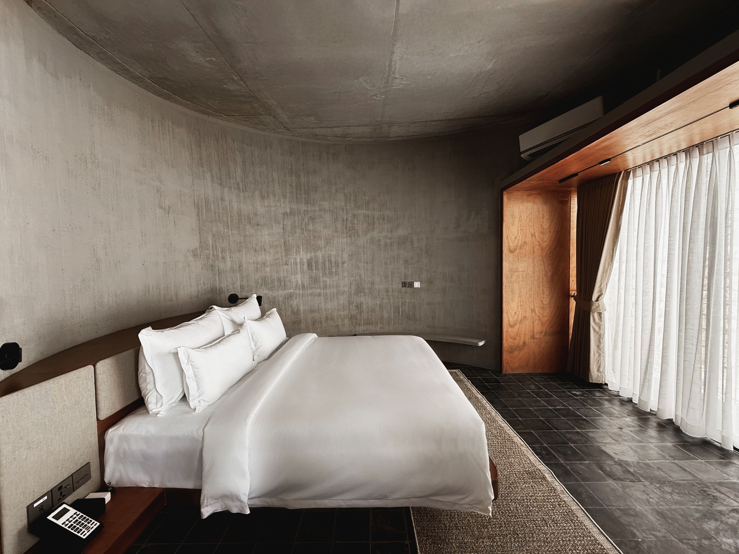 Quarto moderno com uma cama grande, cabeceira de madeira, teto de concreto e vista para uma porta de madeira.